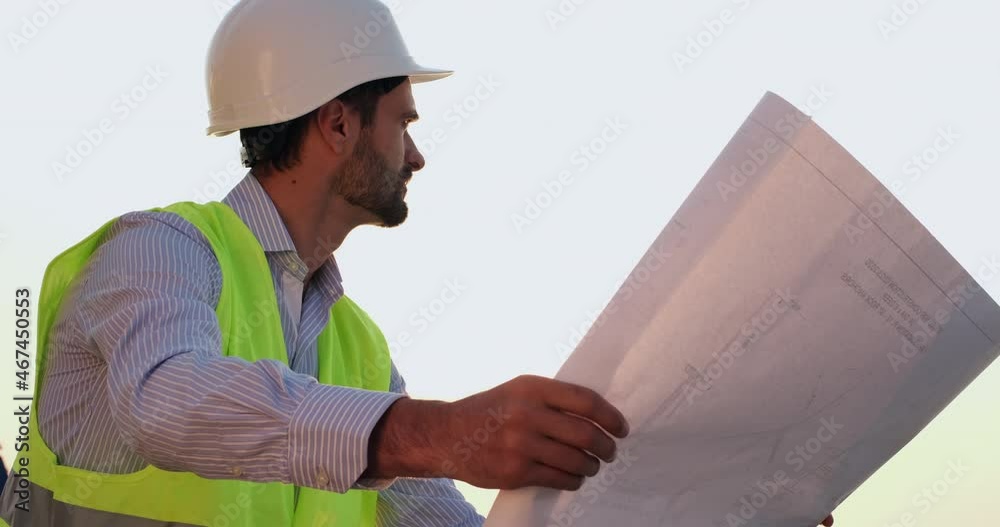Brunet engineer with stubble wearing helmet and vest looks attentively ...
