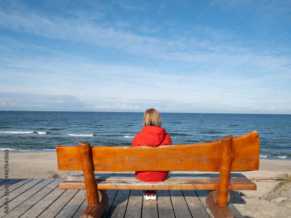 Woman in red coat sits on the bench faced to the sea with waves and ...