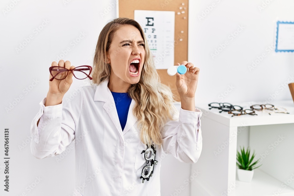 Young beautiful optician woman holding glasses and contact lenses angry ...