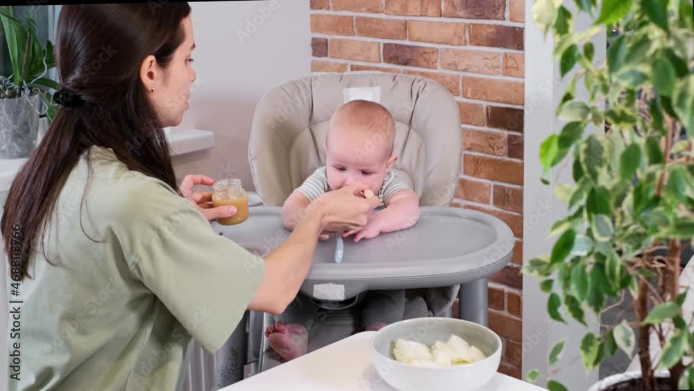 Mother giving food to baby boy, pure from spoon in highchair at home ...