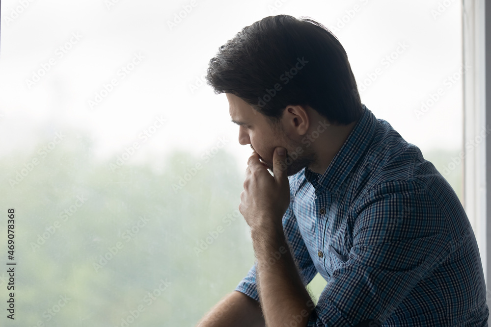 Thoughtful depressed young man touching chin, looking out of window ...