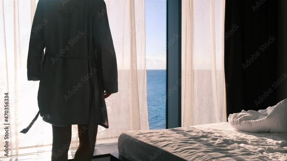 Blonde in a gray silk bathrobe, opens curtains on the window, view of ...