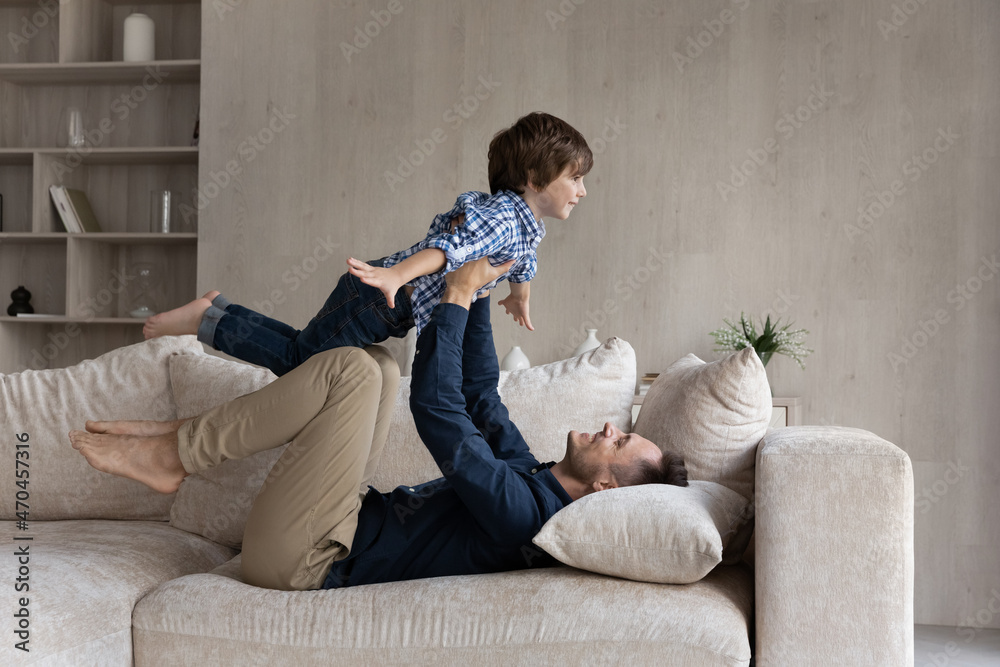 Dad lying on couch lifts on outstretched hands little 6s son. Happy ...