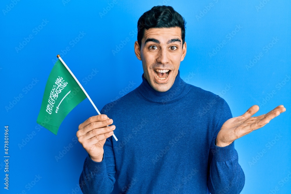 Handsome hispanic man holding kingdom of saudi arabia flag celebrating ...