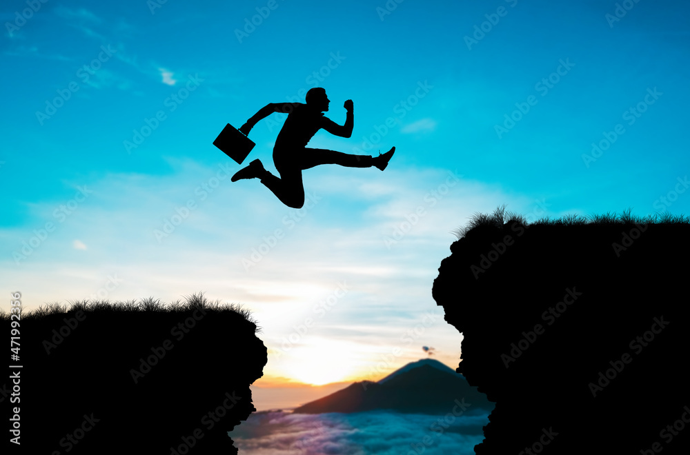 Silhouette of a businessman with a briefcase in his hands, jumping over the abyss between the ...