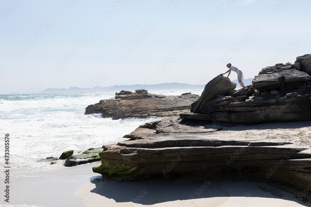 Teenage girl climbing over smooth flat layered rocks on a beach, waves ...