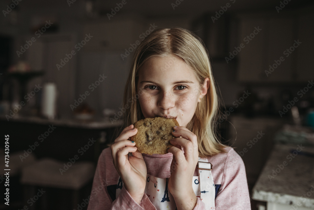 Tween Girl Eating A Large Chocolate Chip Cookie In Kitchen With Apron