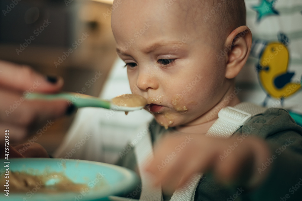 Baby eating with a face stained in food while mother feeds him with a ...