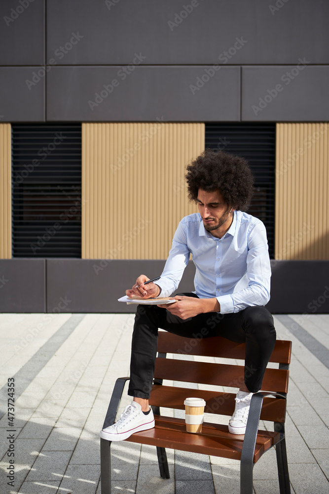 Man with coffee cup writing in book while sitting on chair during sunny ...