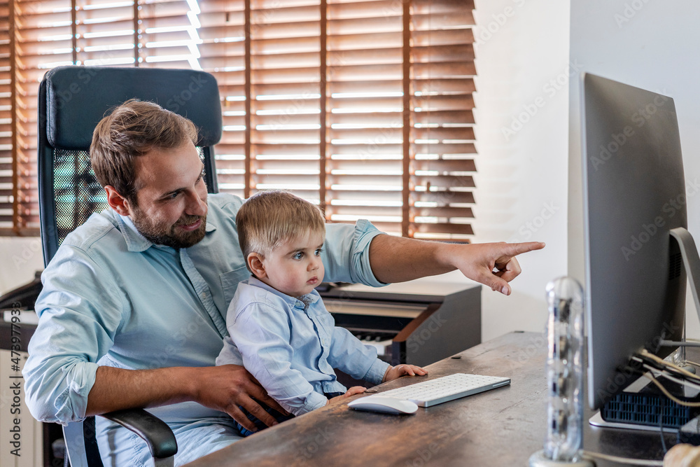 Smiling father pointing at computer while son sitting on lap at home ...