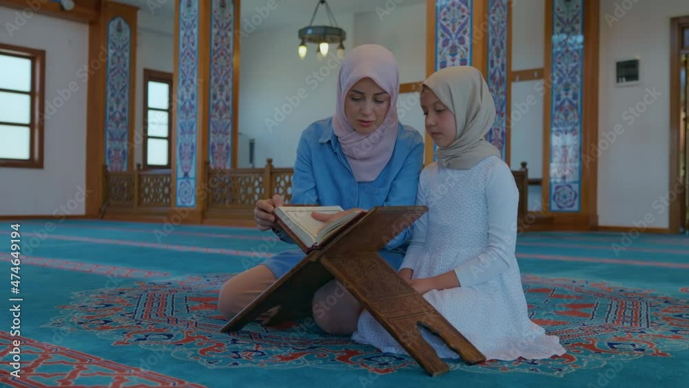 A Muslim mother in a hijab teaches her little daughter to read the Qur ...