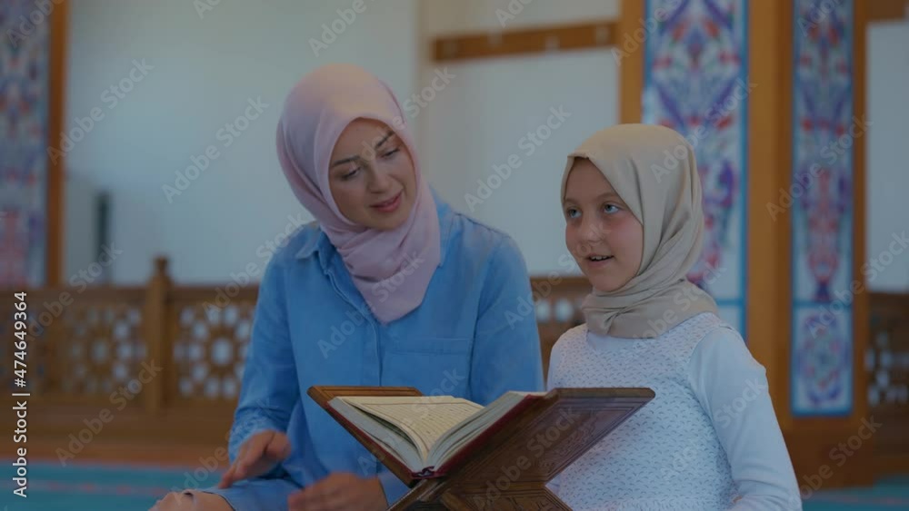 A Muslim mother in a hijab teaches her little daughter to read the Qur'an in a mosque. Little ...
