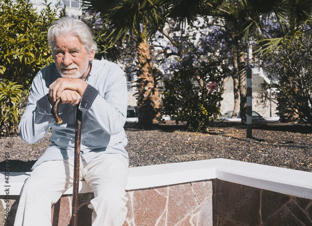 Old senior man with hands over the cane sitting in public park with sad ...