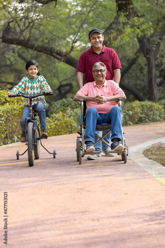 Man pushing old father on wheelchair while his son is riding bicycle at ...