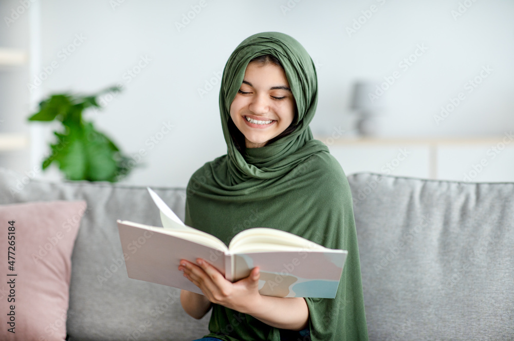 Smiling Islamic teen girl in hijab reading book, enjoying interesting ...