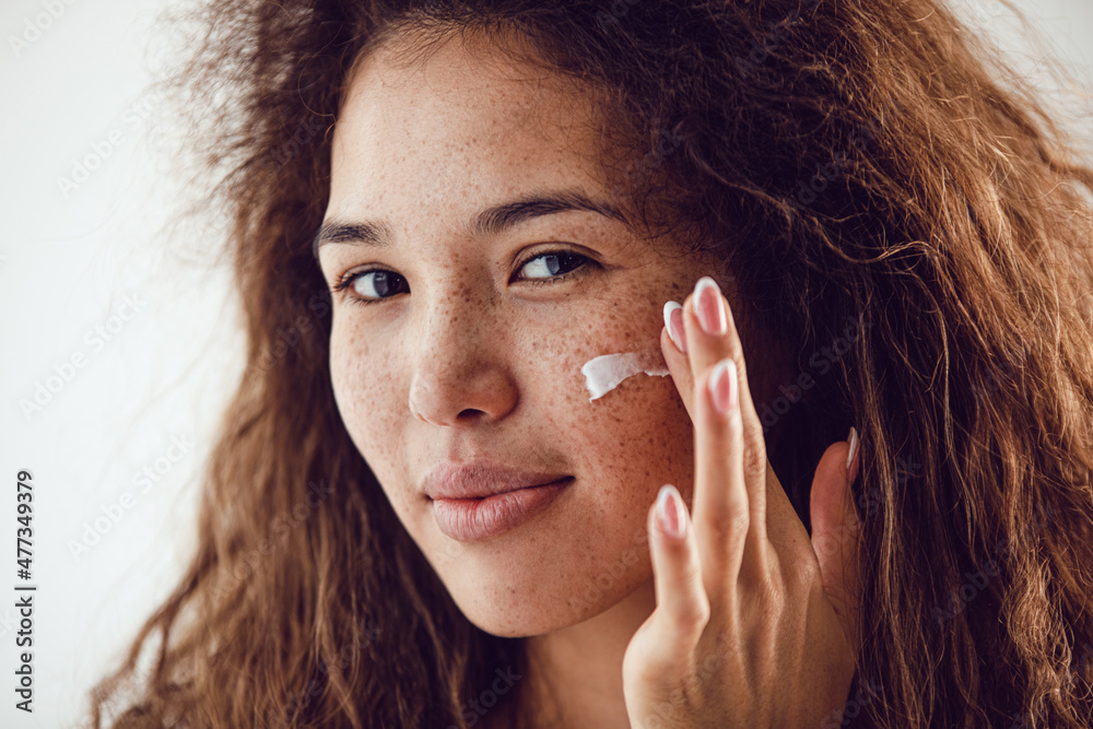 Portrait of woman with curly hair and freckles applying moisturizer to ...