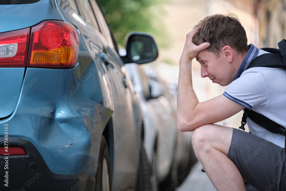 Sad young man driver sitting near his dented car looking shocked on ...