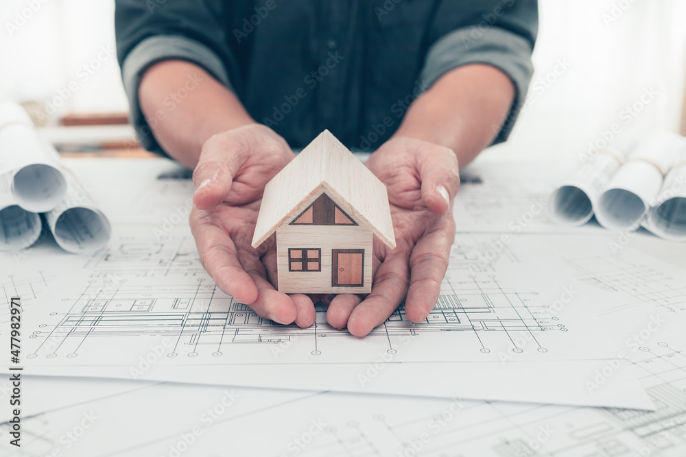 Architect engineer hands hold and show mini house model and use pen drawing on bueprint. House ...