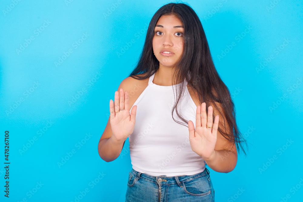 Young hispanic girl wearing tank top over blue background doing stop ...