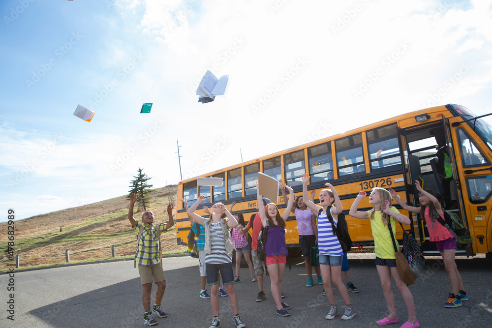Excited children throwing books up in the air by school bus on field ...