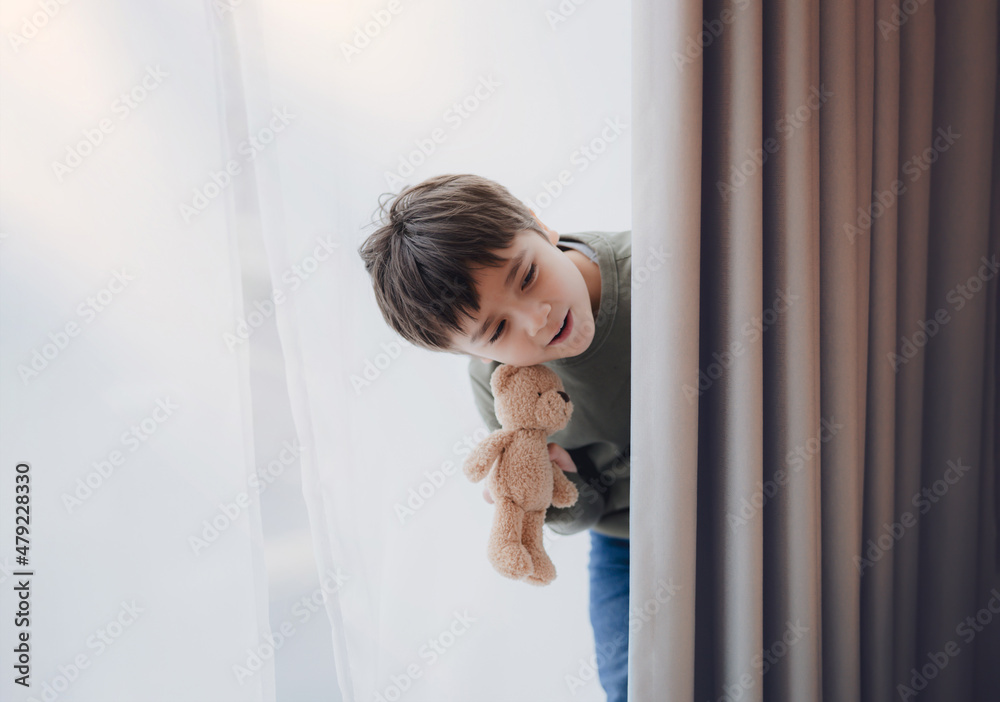 Cute young boy holding teddy bear standing behind lace curtain with ...