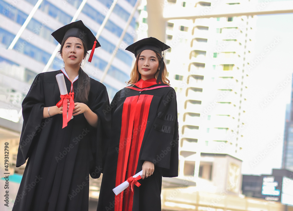 Two happy graduated women in graduation gowns holding diploma, standing ...