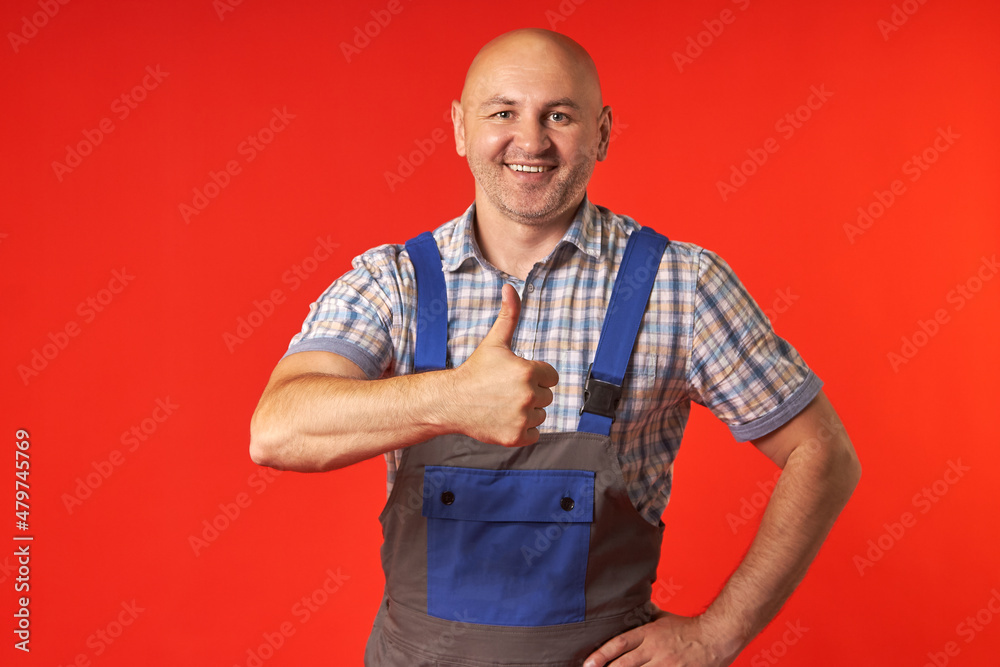 Bald man in overalls and a plaid shirt stands on a red background and ...