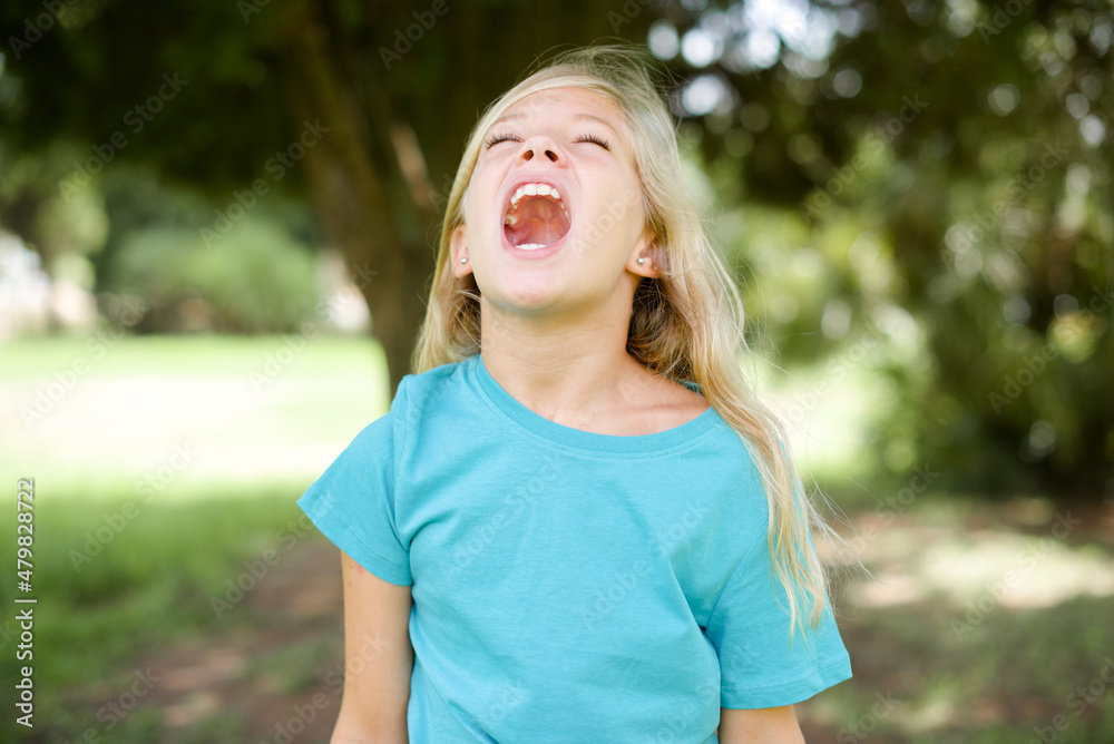 Caucasian little kid girl wearing blue T-shirt standing outdoors angry ...