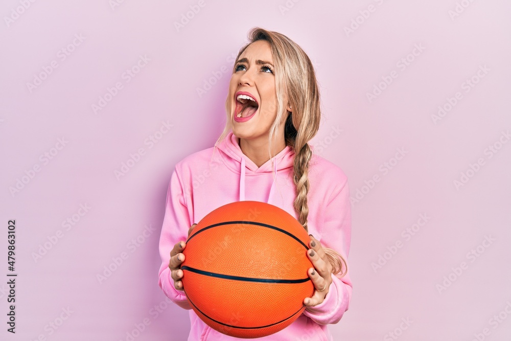 Beautiful young blonde woman holding basketball ball angry and mad ...