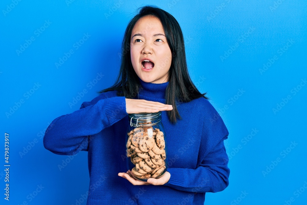 Young chinese girl holding jar of chocolate chips cookies angry and mad ...