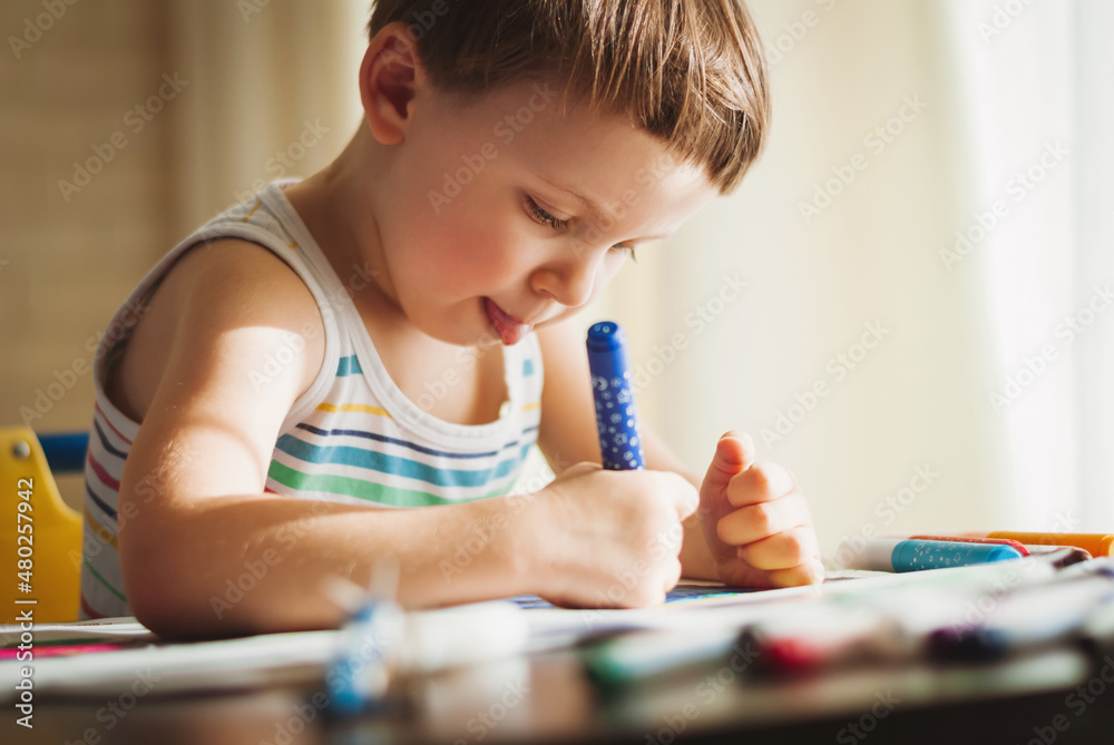 The boy is sitting at the table and diligently draws something with ...