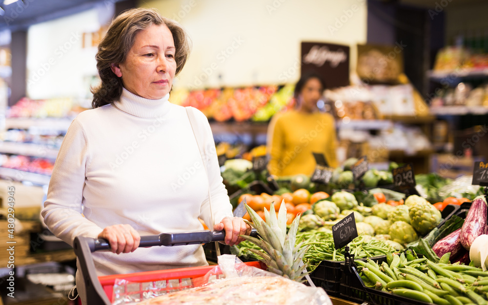 Pensive middle aged female shopping at store, walking among shelves and ...
