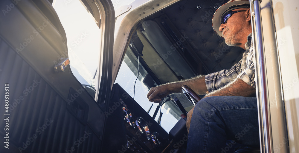 Elderly Latino semi truck driver sits in the cab looking ahead wearing ...