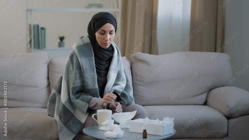 Sick young woman in black hijab wrapped herself in blanket sit on couch ...