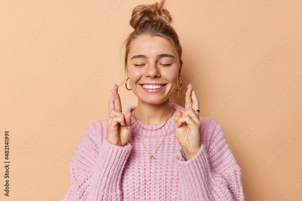 Positive hopeful young womam keeps eyes closed smiles broadly crosses ...