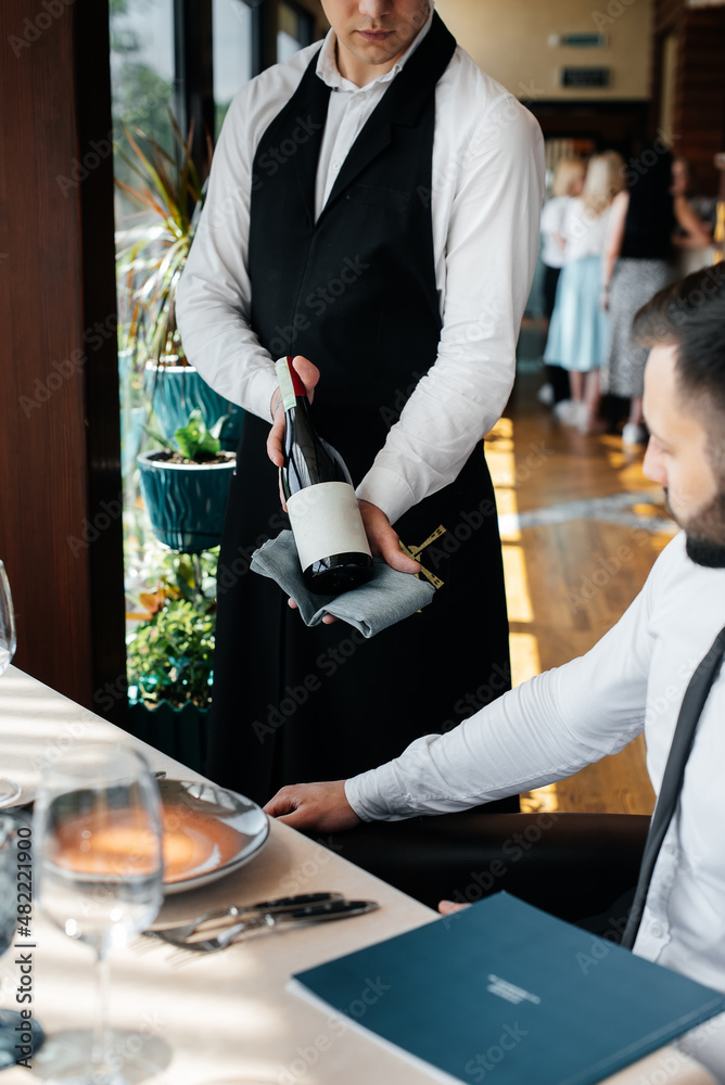 A young sommelier in a stylish uniform demonstrates and offers the ...
