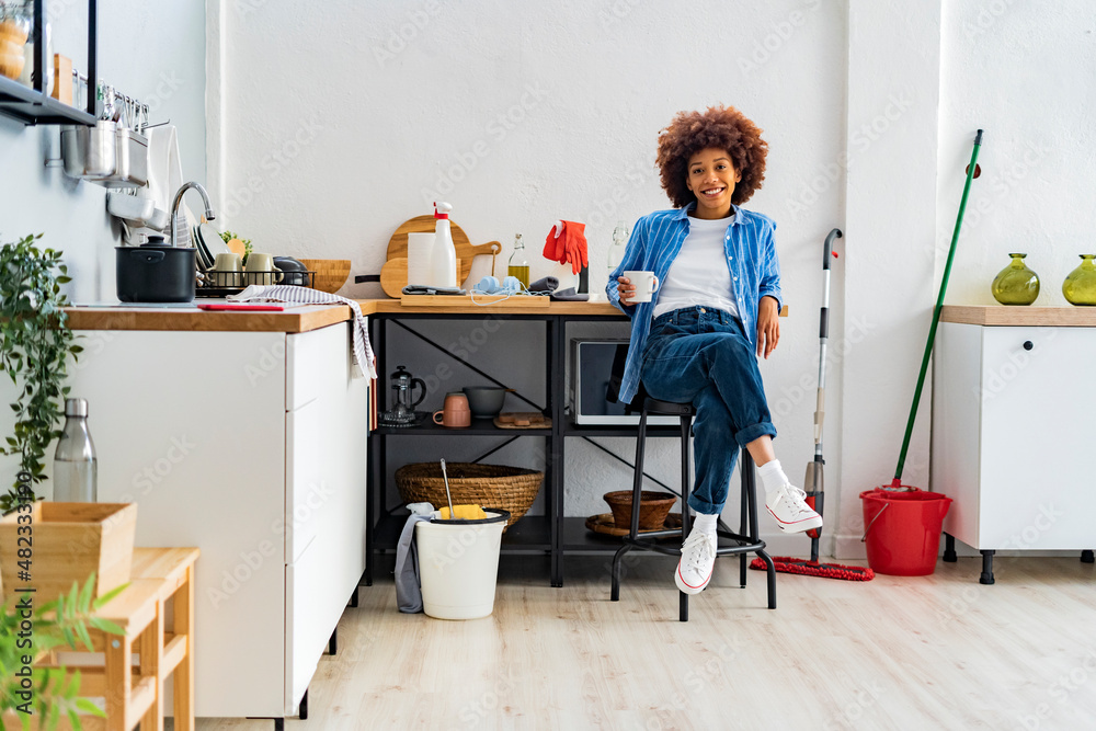Smiling young Afro woman sitting on stool leaning on kitchen counter ...