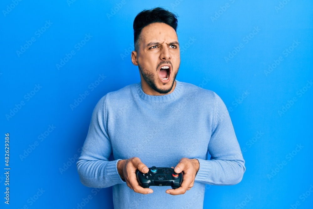 Young arab man playing video game holding controller angry and mad ...