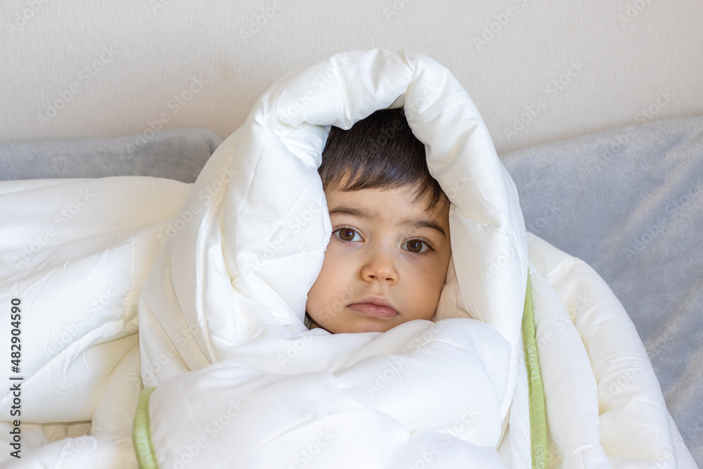 cute 4 years old boy turned wrapped into white soft blanket is making different faces. kid is ...