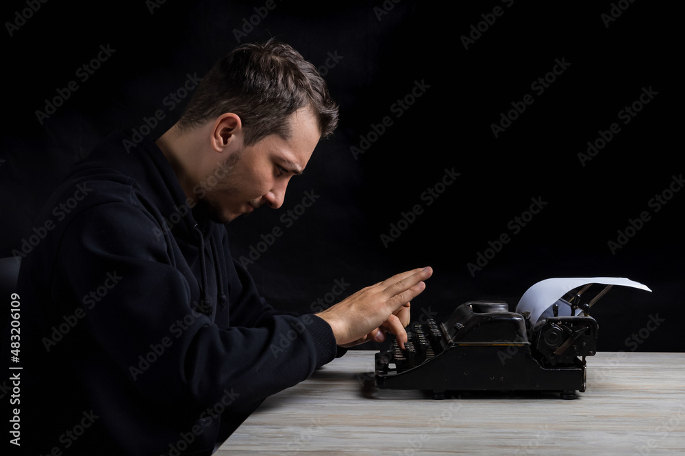 Young handsome man in black hoodie is typing on a typewriter, on black ...