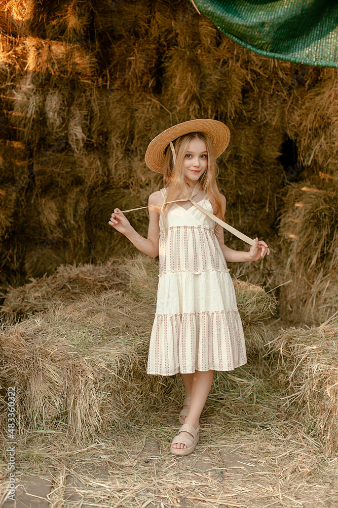 Romantic tween girl in light sundress and straw hat standing on hayloft ...