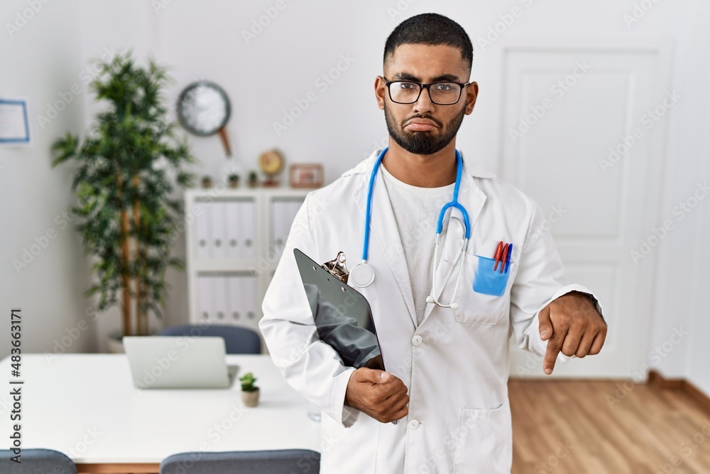 Young indian man wearing doctor uniform and stethoscope pointing down ...