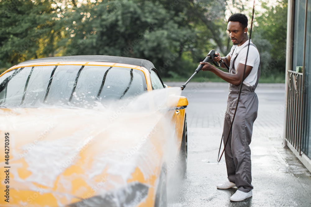 Handsome African guy in t-shirt and gray overalls washing his yellow ...
