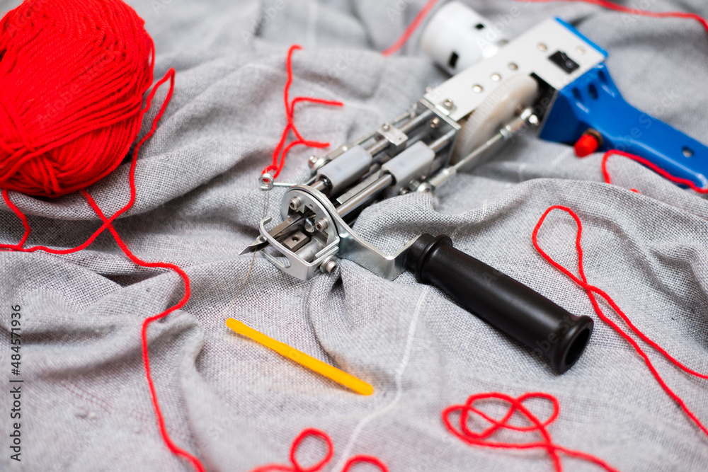 The tufting gun lies on a gray burlap, ready to work with the red yarn ...