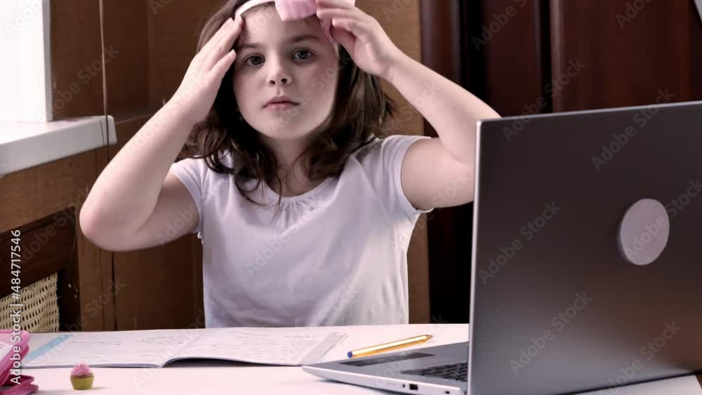 A little schoolgirl girl sits at a table with a laptop does her ...