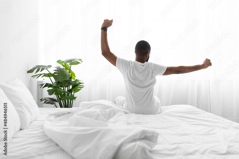Young black man in white clothes sits on bed, stretching his body and ...
