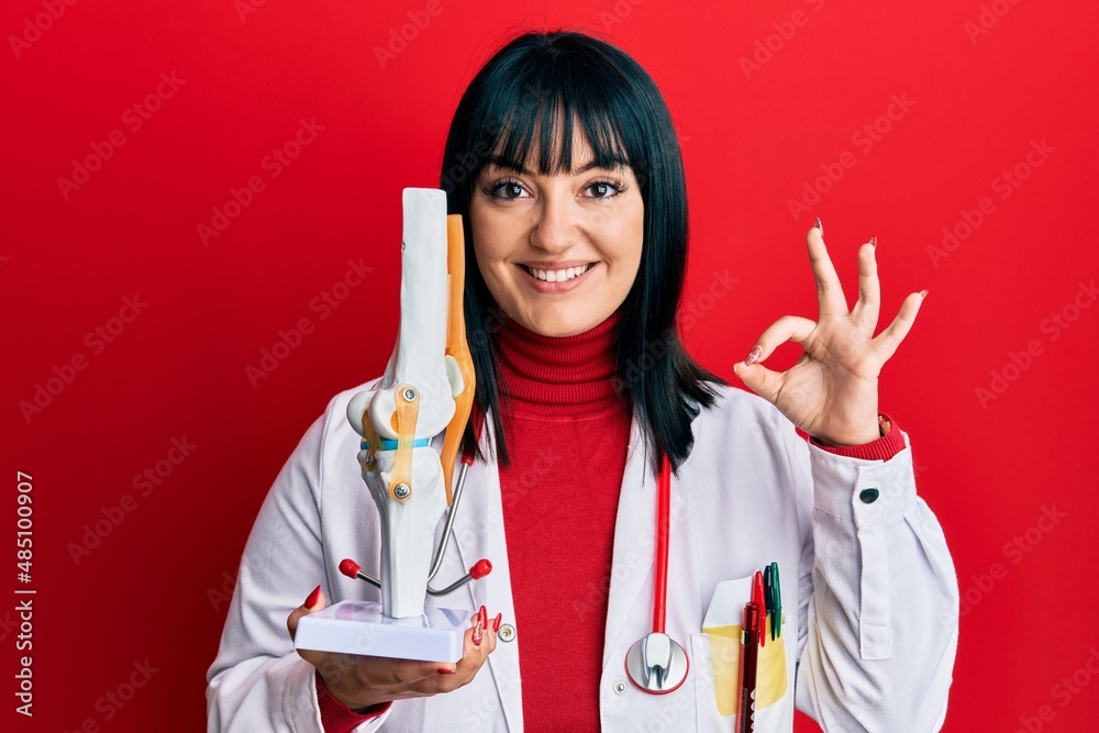 Young hispanic doctor woman holding anatomical model of knee joint ...