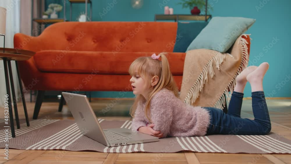 Little school kid girl use laptop computer lying on floor alone at home ...