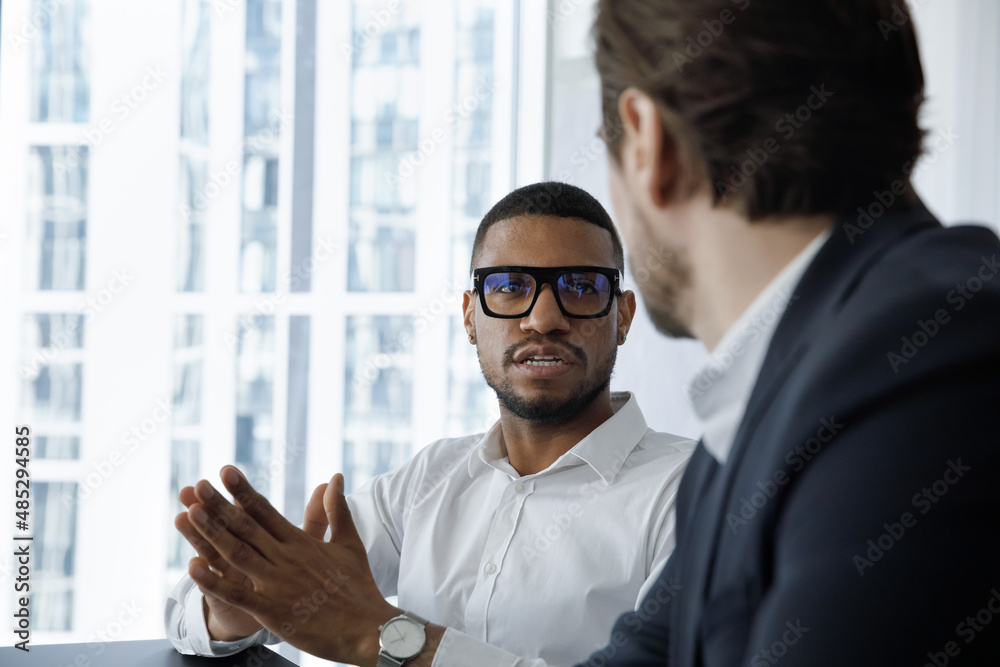Serious engaged African employee in glasses talking to coworker ...