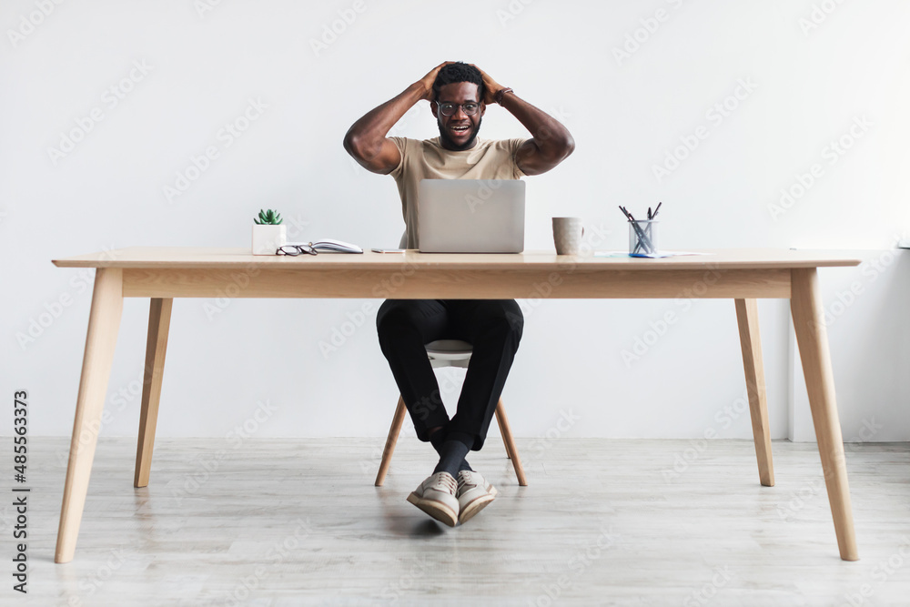 Shocked black man sitting at desk with laptop, grabbing head in terror ...